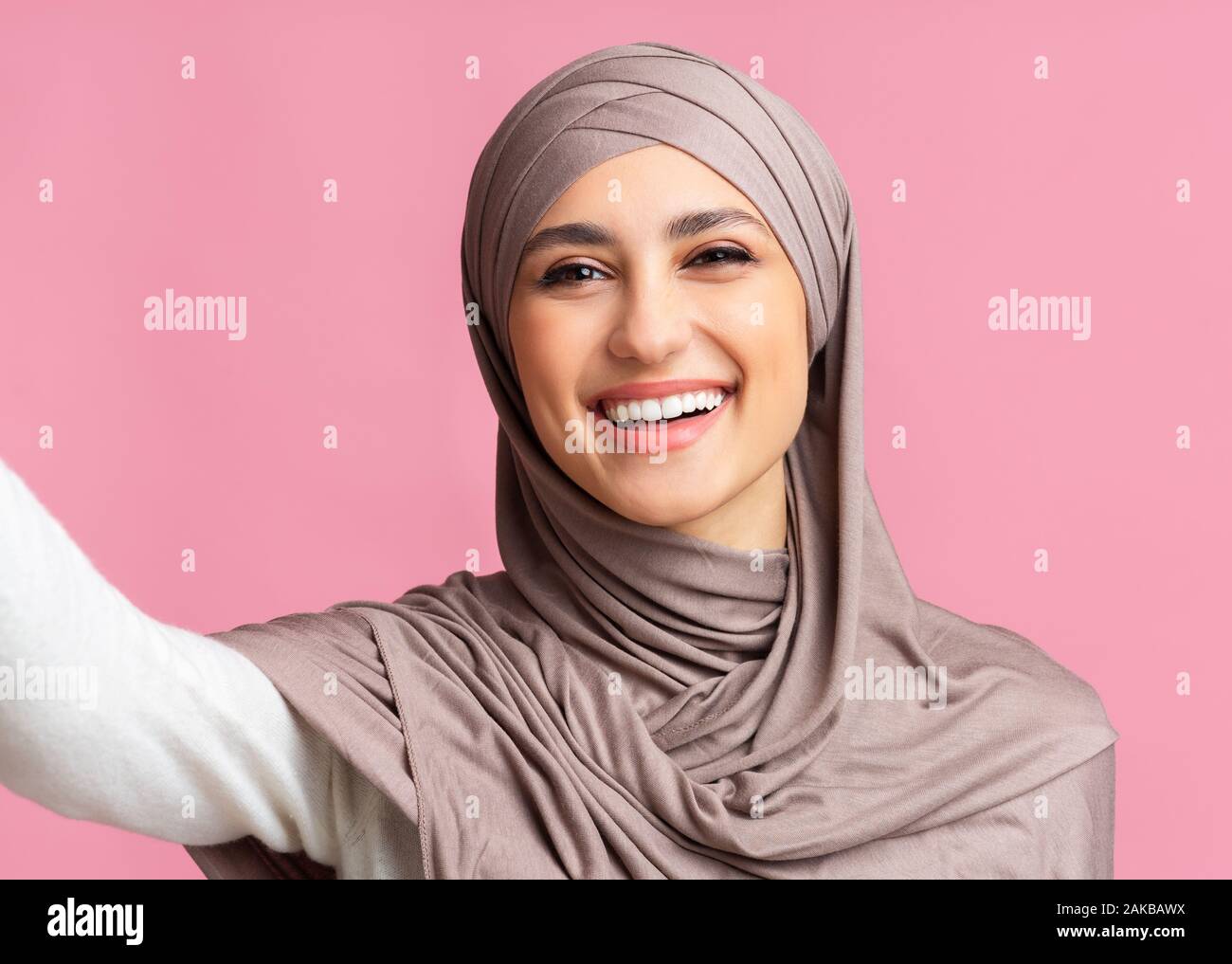 Cheerful arabic girl in hijab taking selfie and sincerely laughing, pink background with free ...