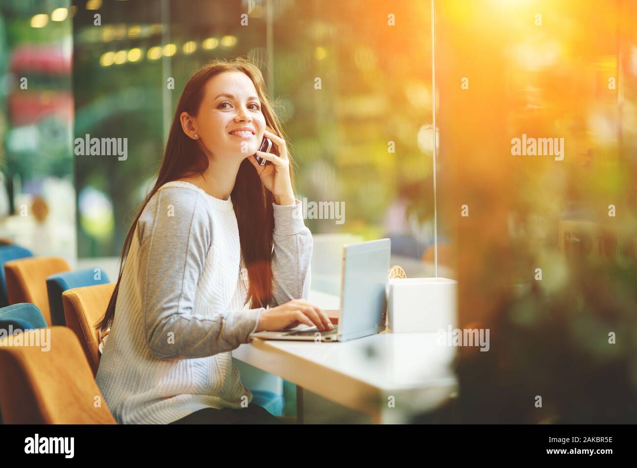 Beautiful Caucasian woman uses Phone while sitting with portable net-book in modern cafe bar ...