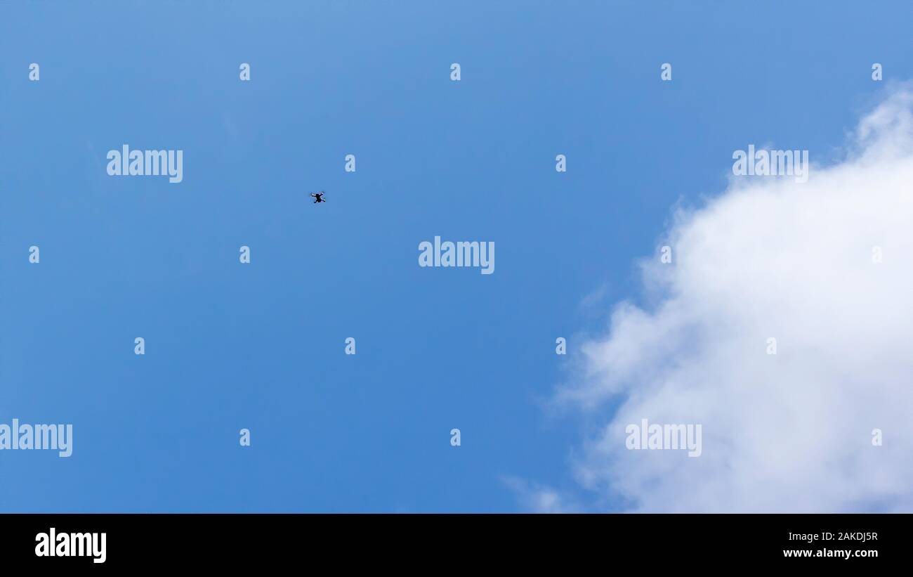 A tiny drone high in the sky overhead, with white clouds in the ...