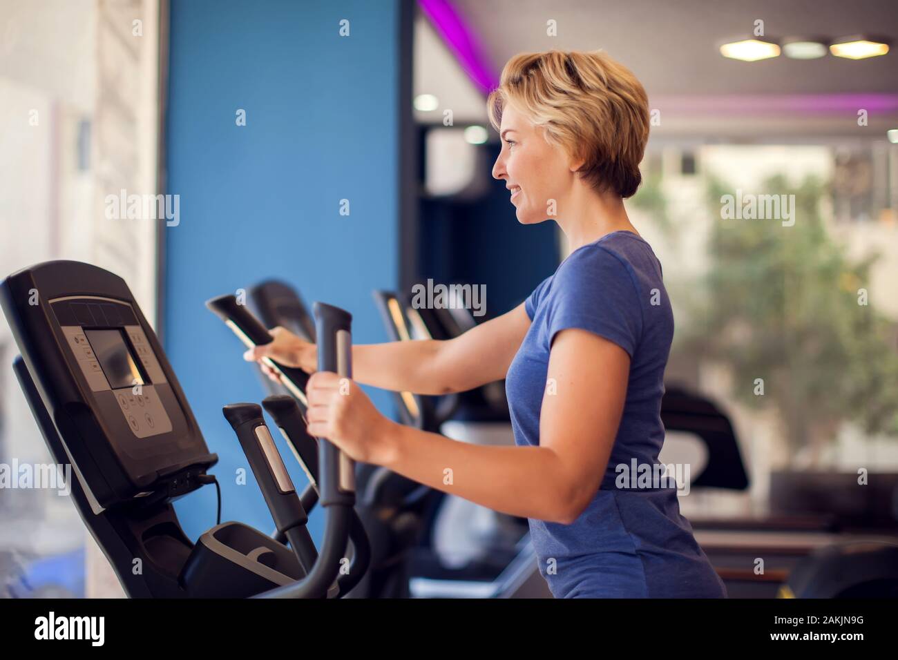 Woman in blue shirt and short hair training in cardio zone in the gym ...