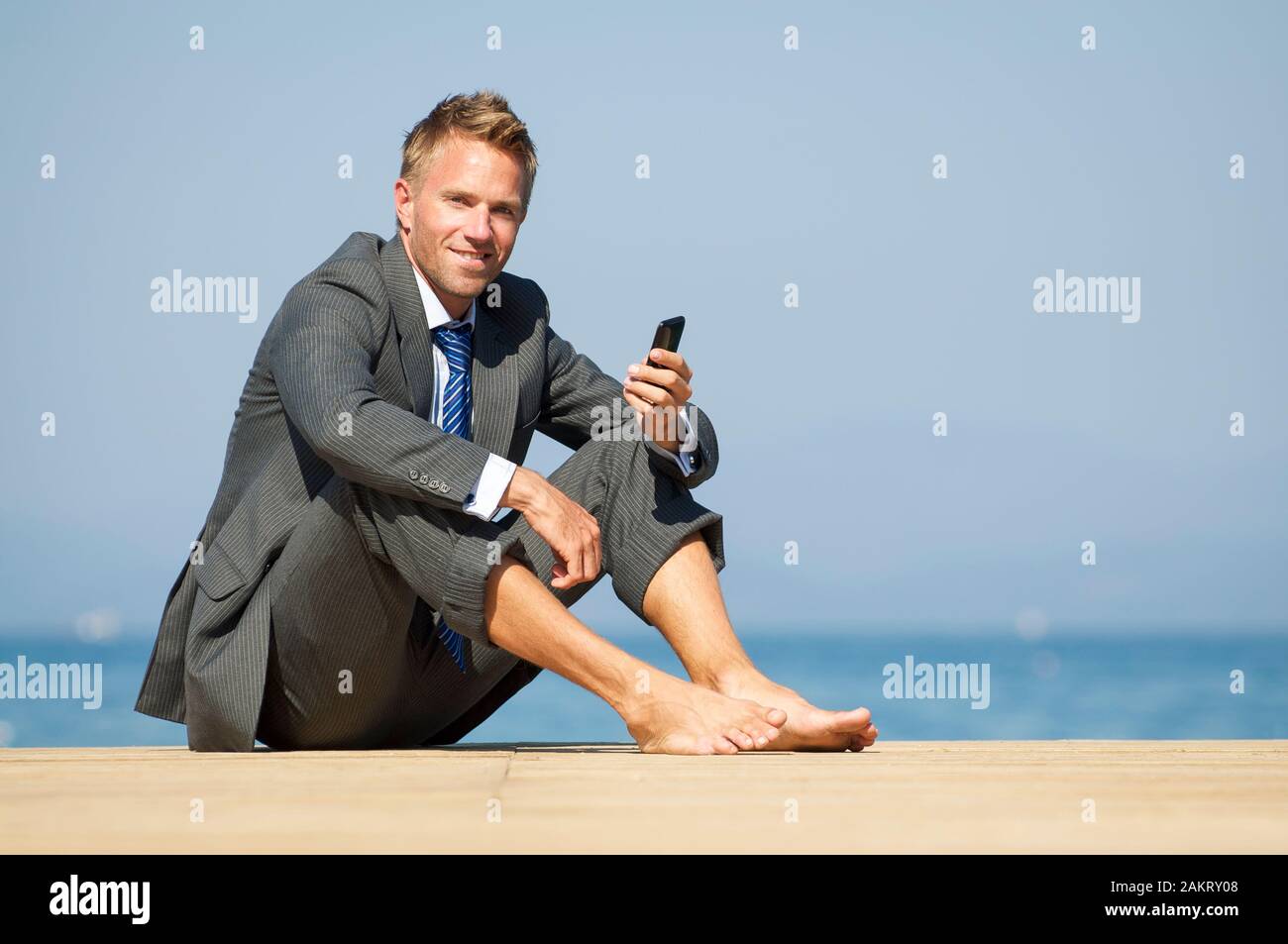 Relaxed barefoot businessman sitting on a sunny dock by the sea using ...