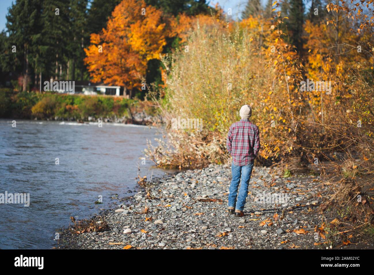 Teen boy in jeans, plaid shirt, and beanie walks along river in fall ...