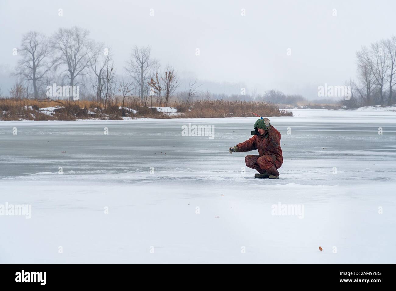 A fisherman sits on the ice of a frozen river and catches fish. Winter ...