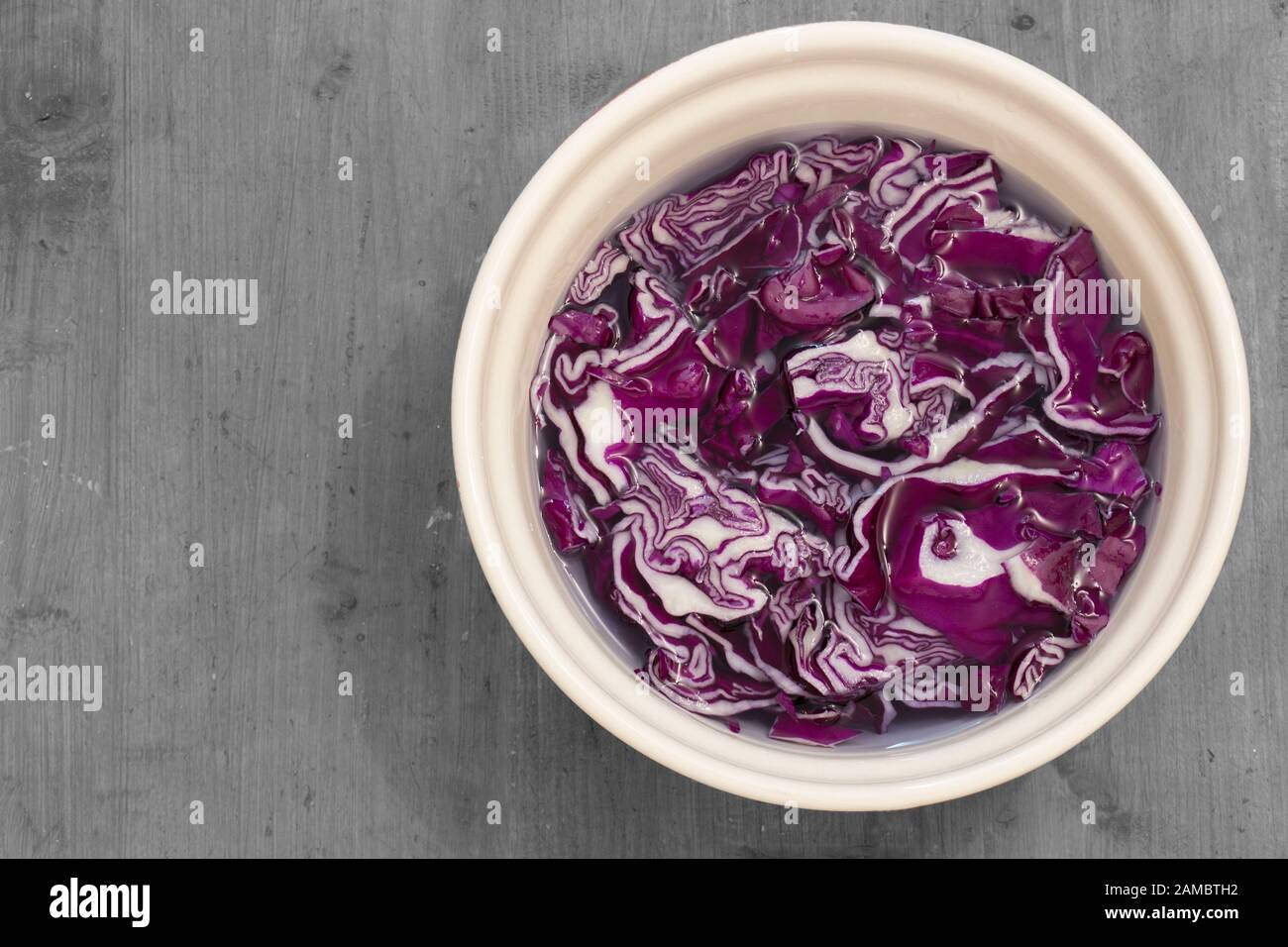 Sliced red cabbage soaking in water in a ceramic bowl. With a grey wood ...