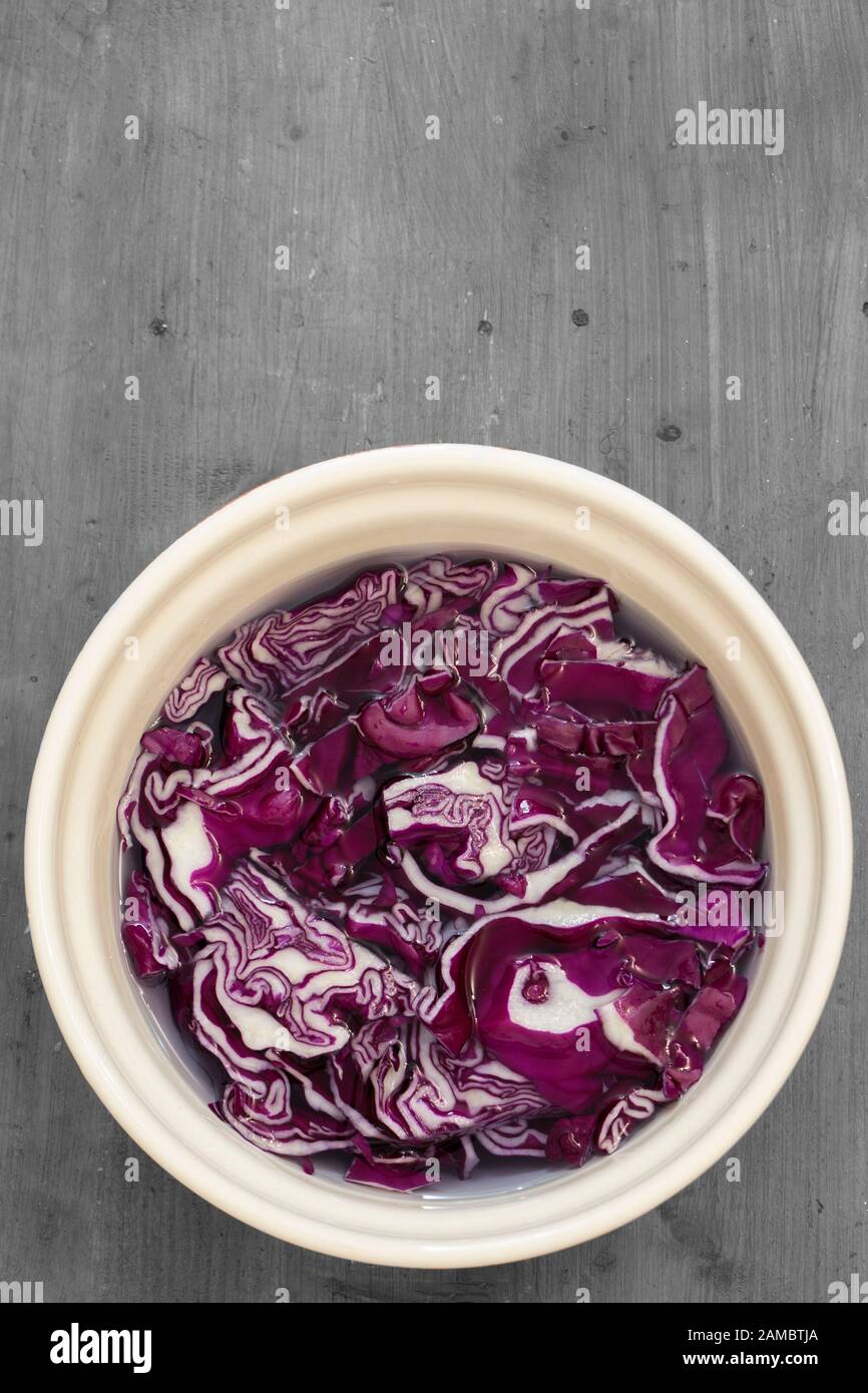 Sliced red cabbage soaking in water in a ceramic bowl. With a grey wood ...