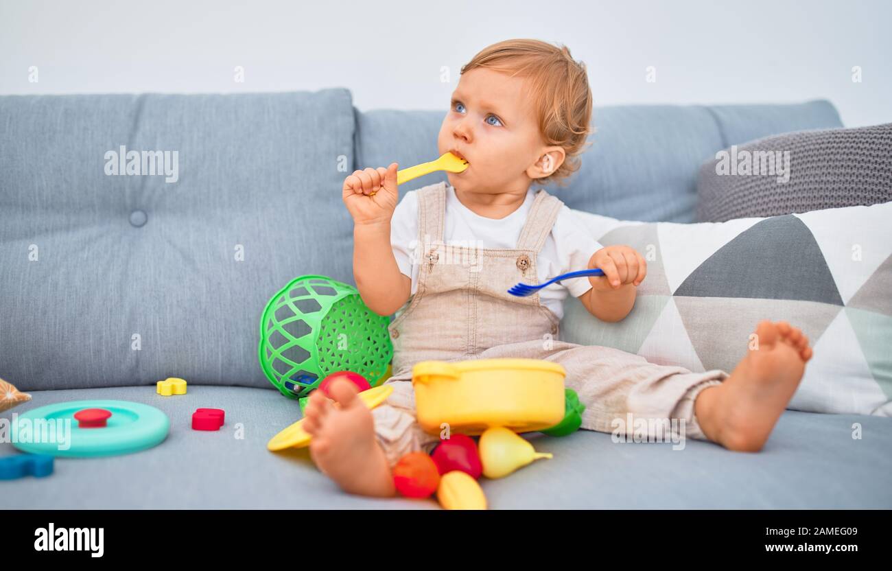 Adorable blonde toddler sitting on the sofa playing with plastic meals ...