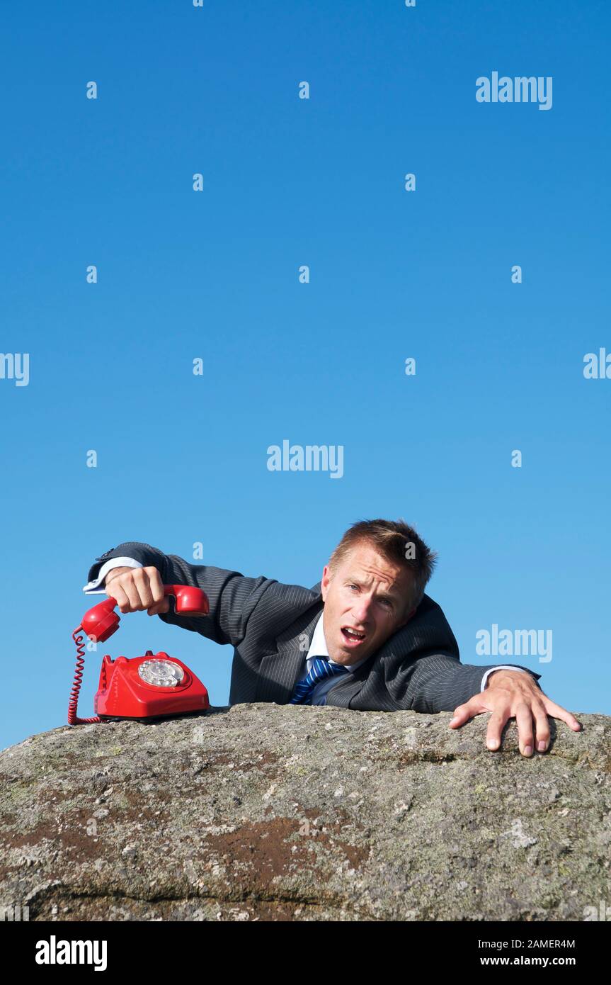 Businessman dangling off a rock cliff reaching to make a desperate call ...