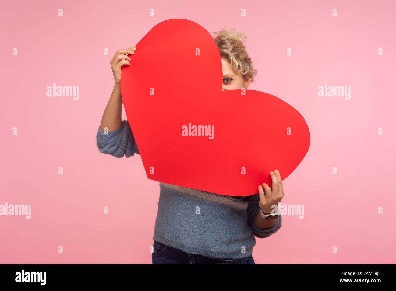 Positive happy woman with curly hair hiding face behind big red heart ...