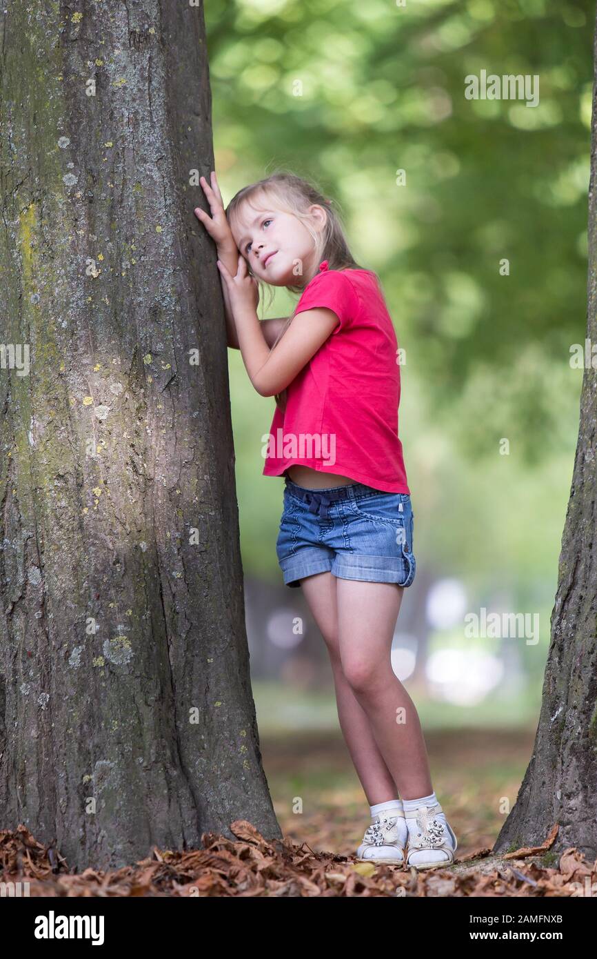 Portrait of a pretty little child girl standing near big tree trunk in ...