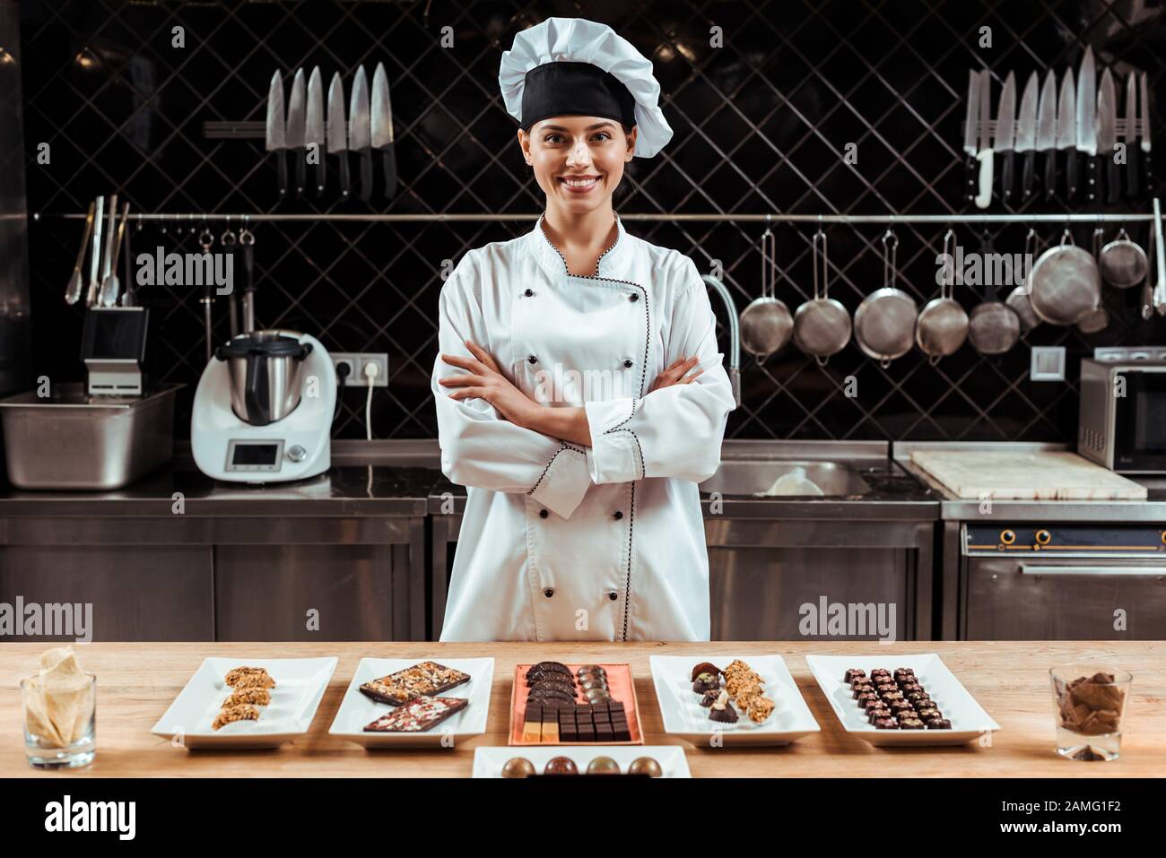 happy chocolatier in chef hat standing with crossed arms near tasty ...