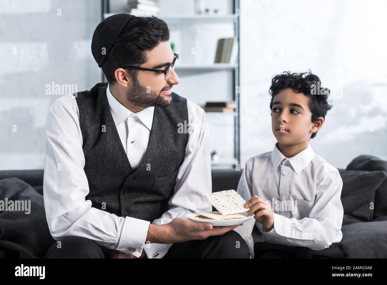 smiling jewish father holding plate and son taking matza in apartment ...