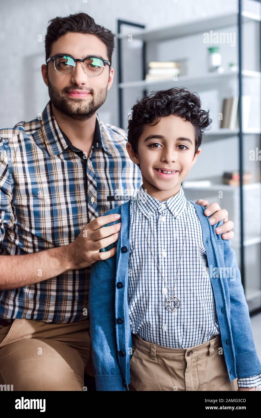 smiling jewish father hugging son and looking at camera in apartment ...