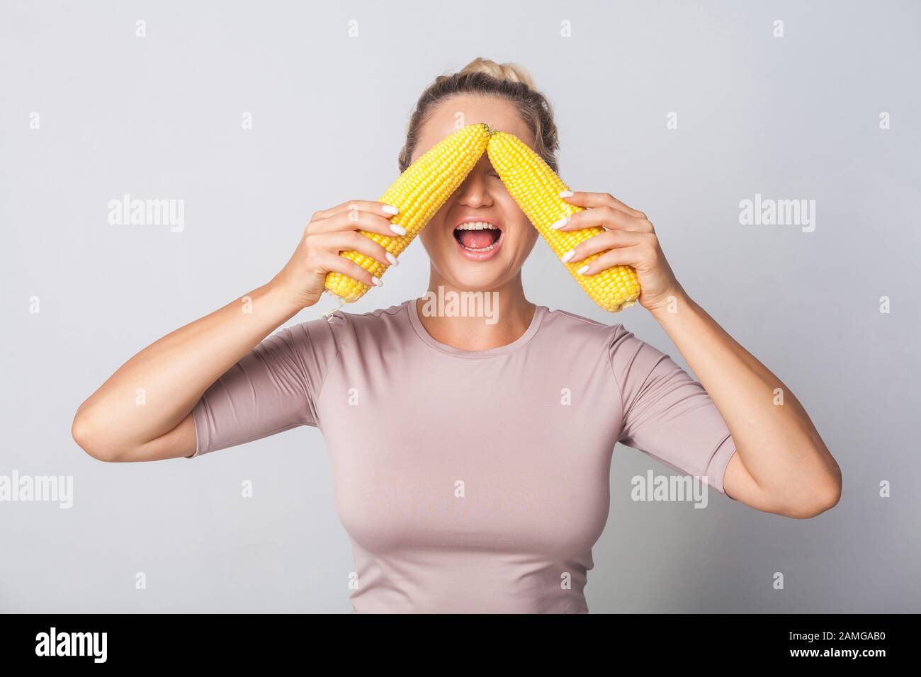 Portrait of cheerful woman covering eyes with corn cobs, opening mouth ...