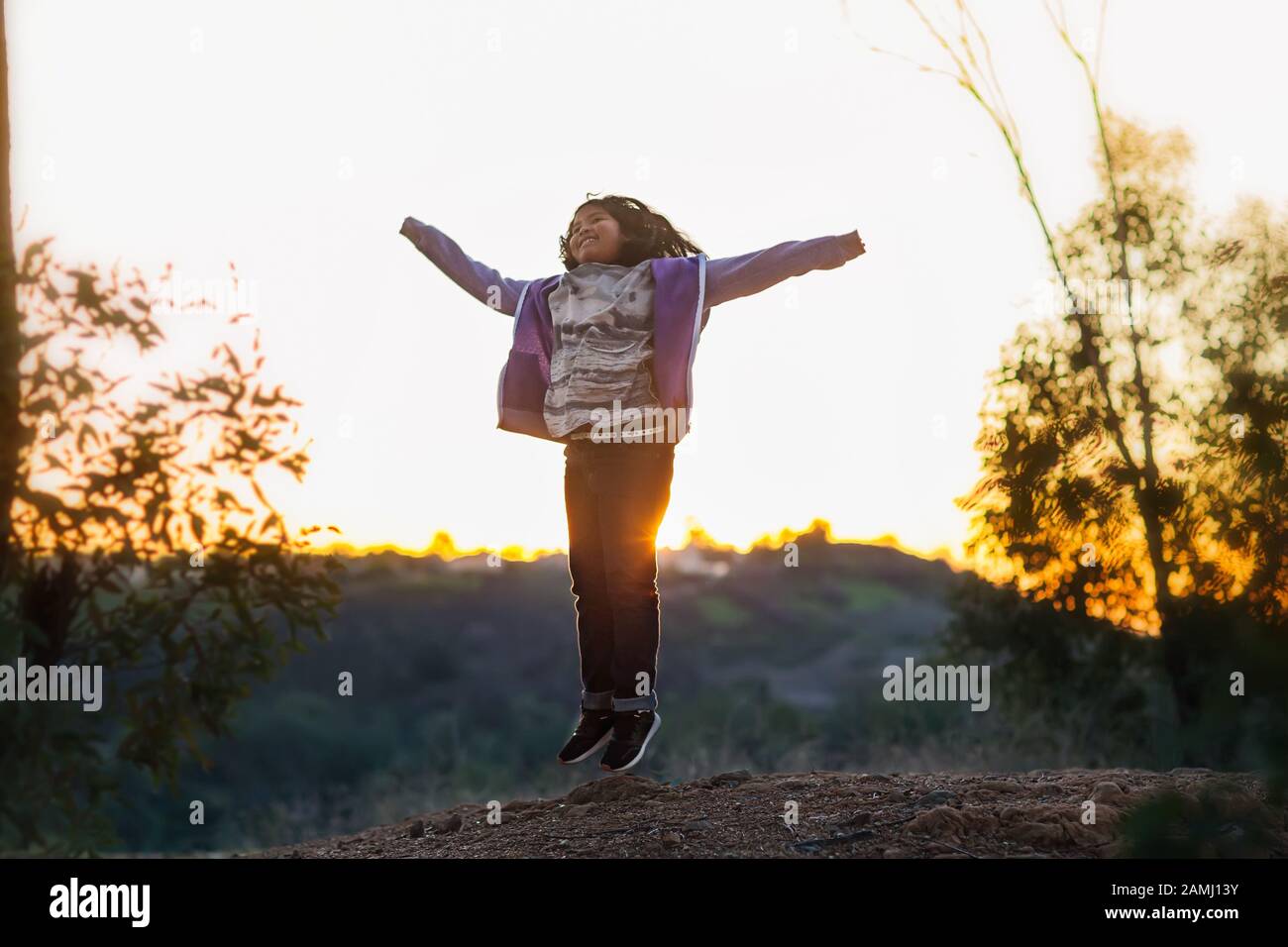 A young girl jumping with arms up on the top of a hill during a sunset ...