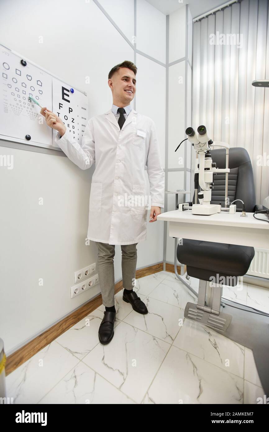 Full length portrait of smiling young ophthalmologist pointing at ...