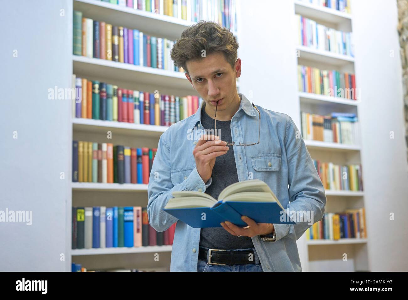 Young, seriously looking man, standing in library with glasses on his ...