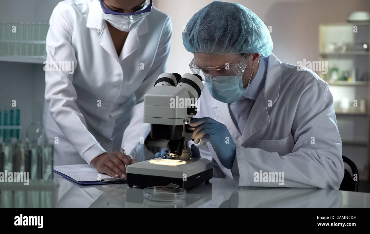 Medical researcher looking through microscope and colleague taking ...