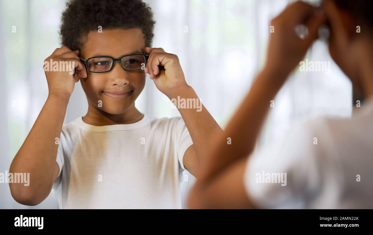 Happy little kid trying on eyeglasses, looking in mirror satisfied good ...
