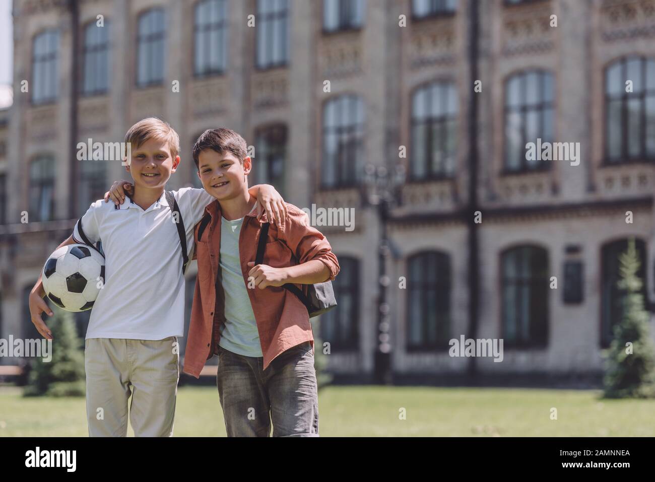 two cheerful schoolboys hugging while standing near school with soccer ...