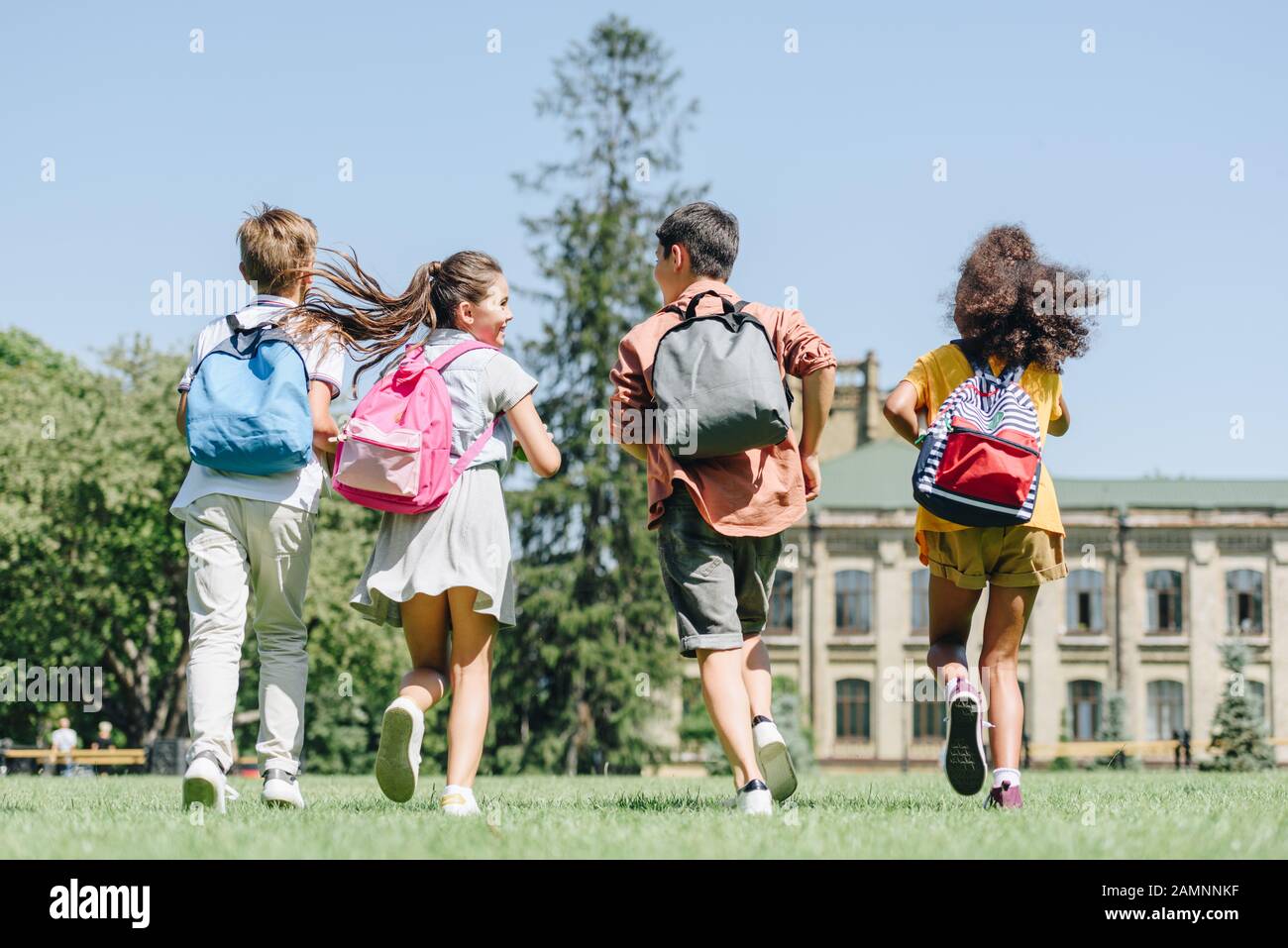 back view of four multiethnic schoolkids with backpacks running on lawn ...