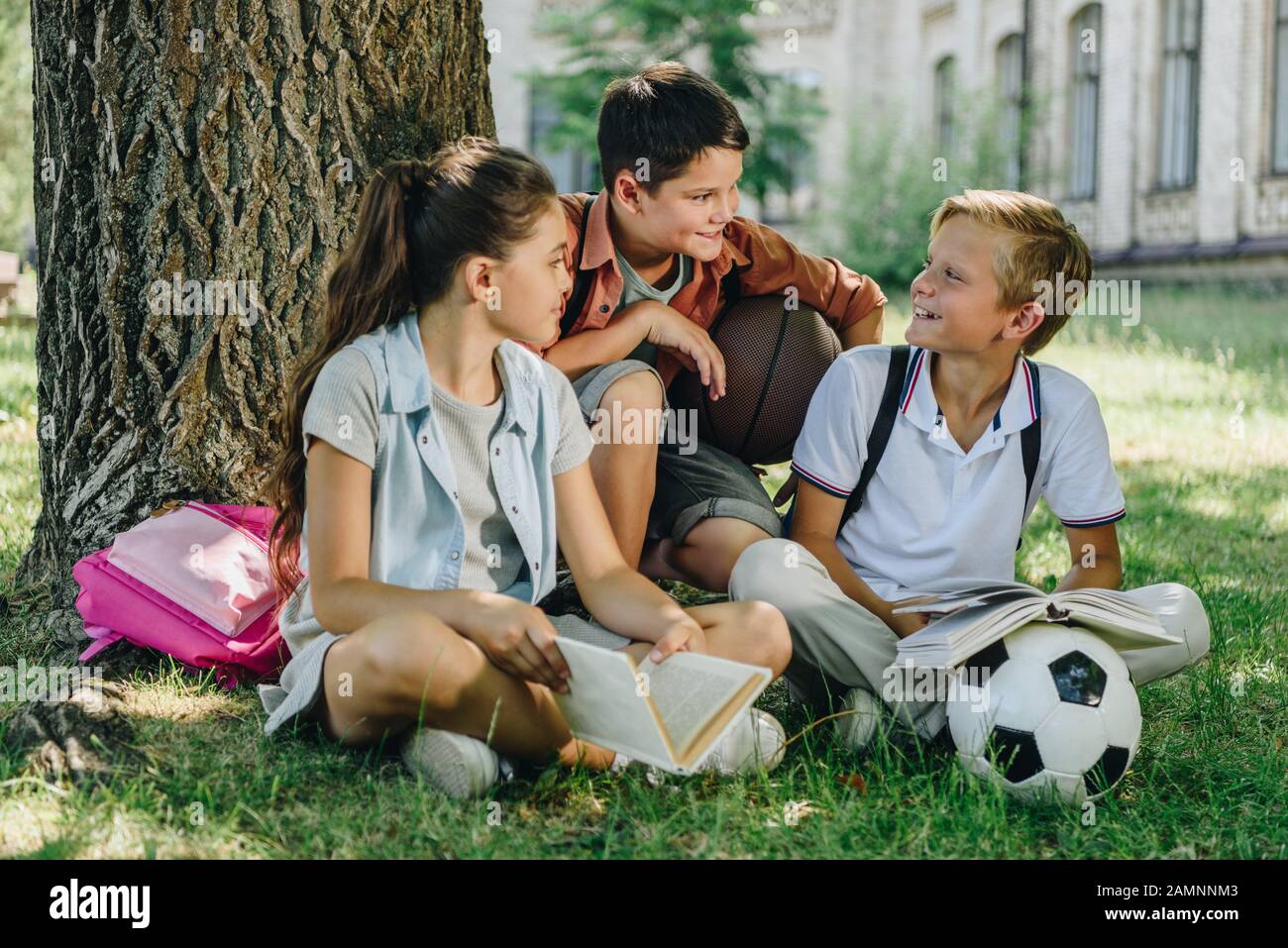 three cheerful schoolkids talking while sitting on lawn under tree ...
