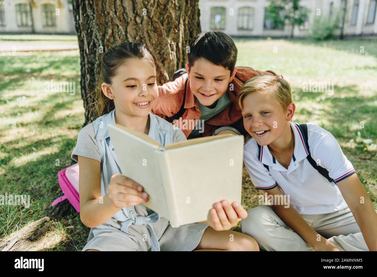 three cheerful schoolkids reading book while sitting under tree in park ...