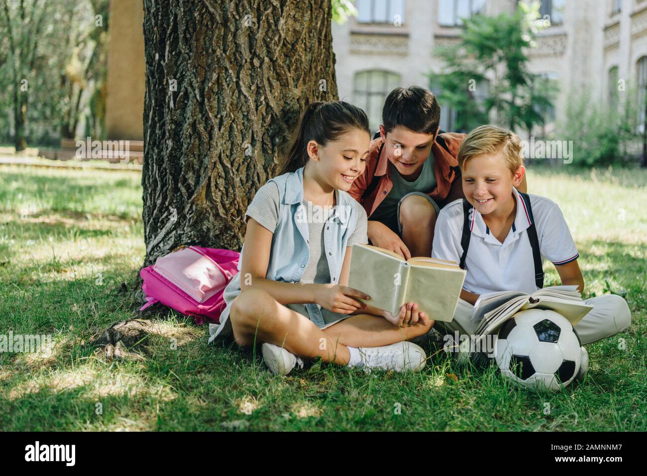 three cute classmates reading book and smiling while sitting on lawn ...