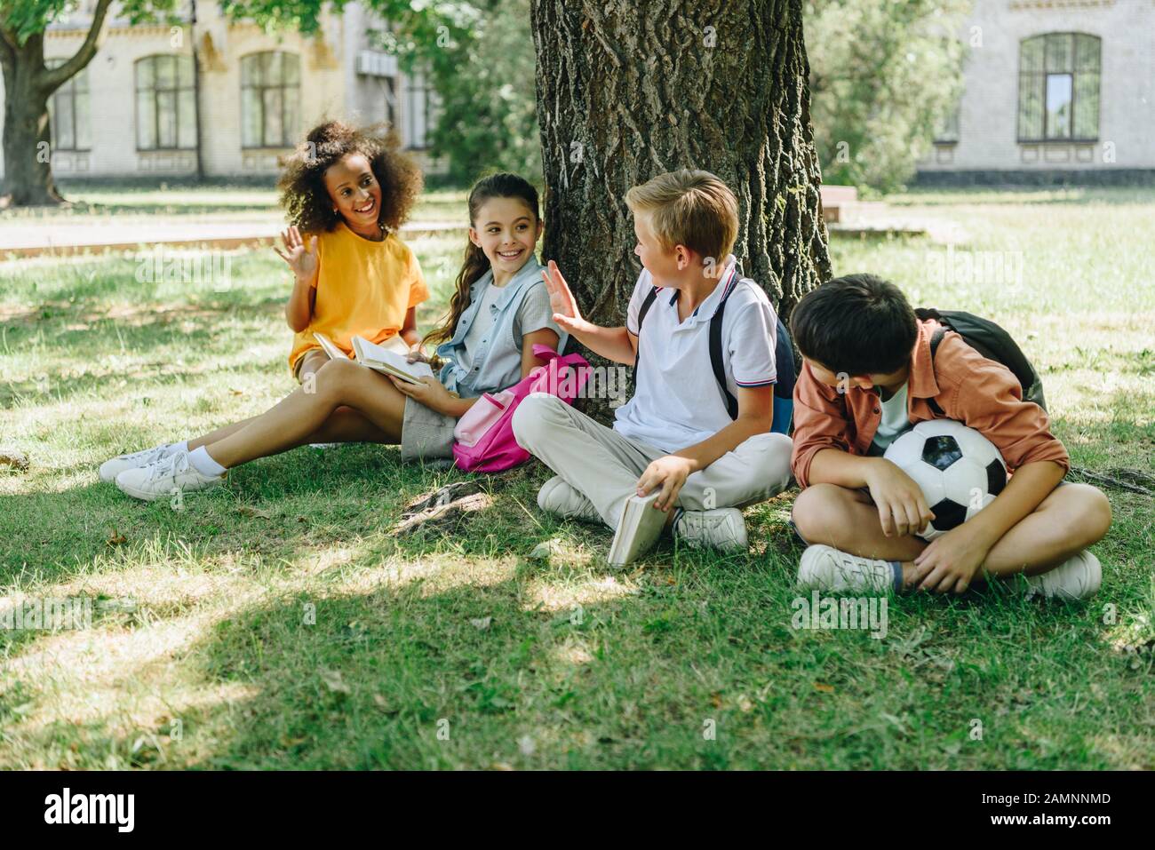four cute multicultural schoolkids talking while sitting on lawn under ...