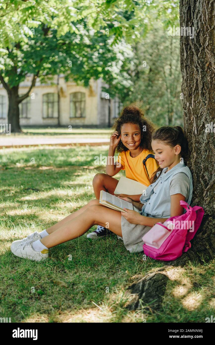 two multicultural schoolgirls reading books while sitting on lawn under ...