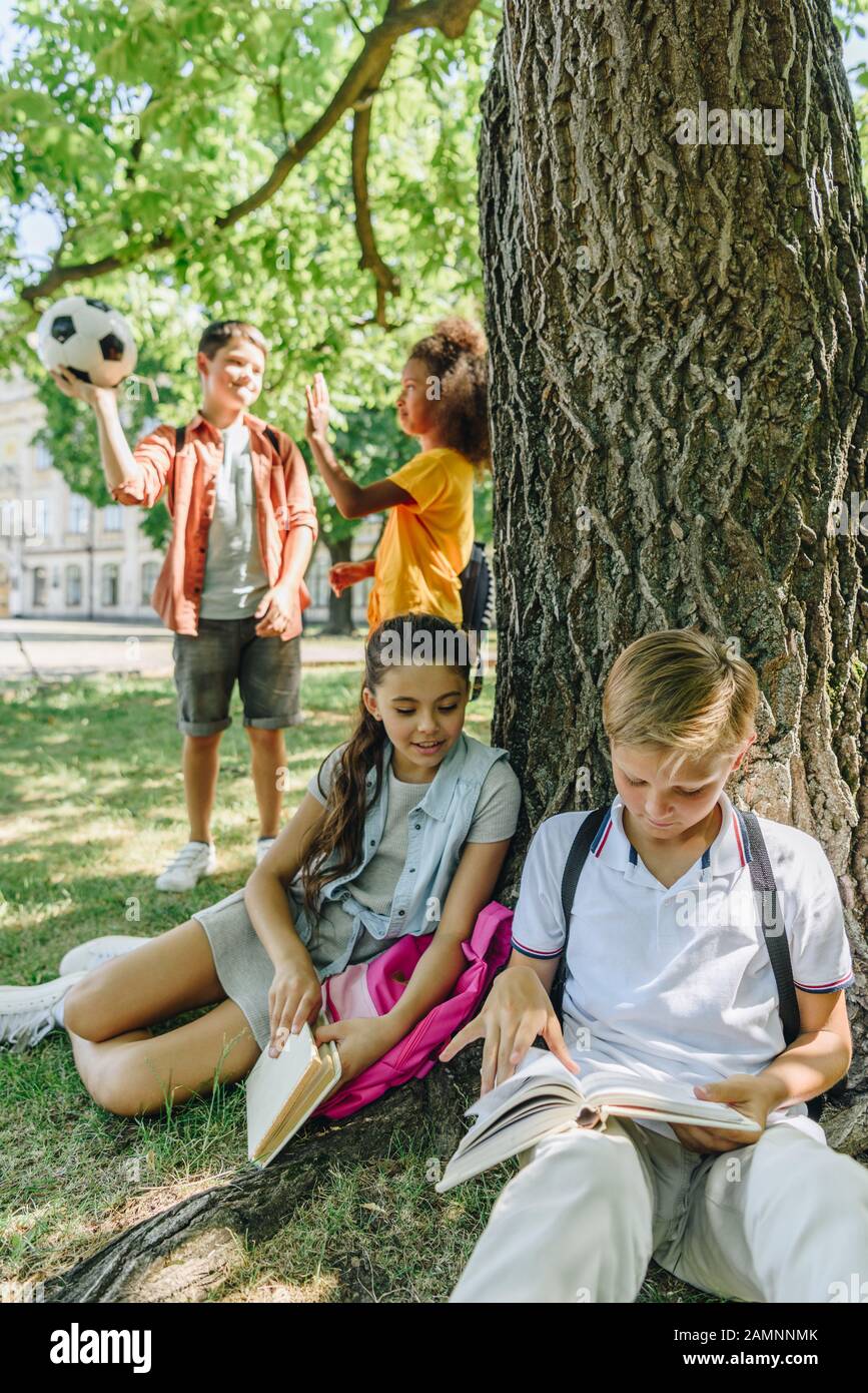 adorable schoolkids sitting under tree and reading books near ...