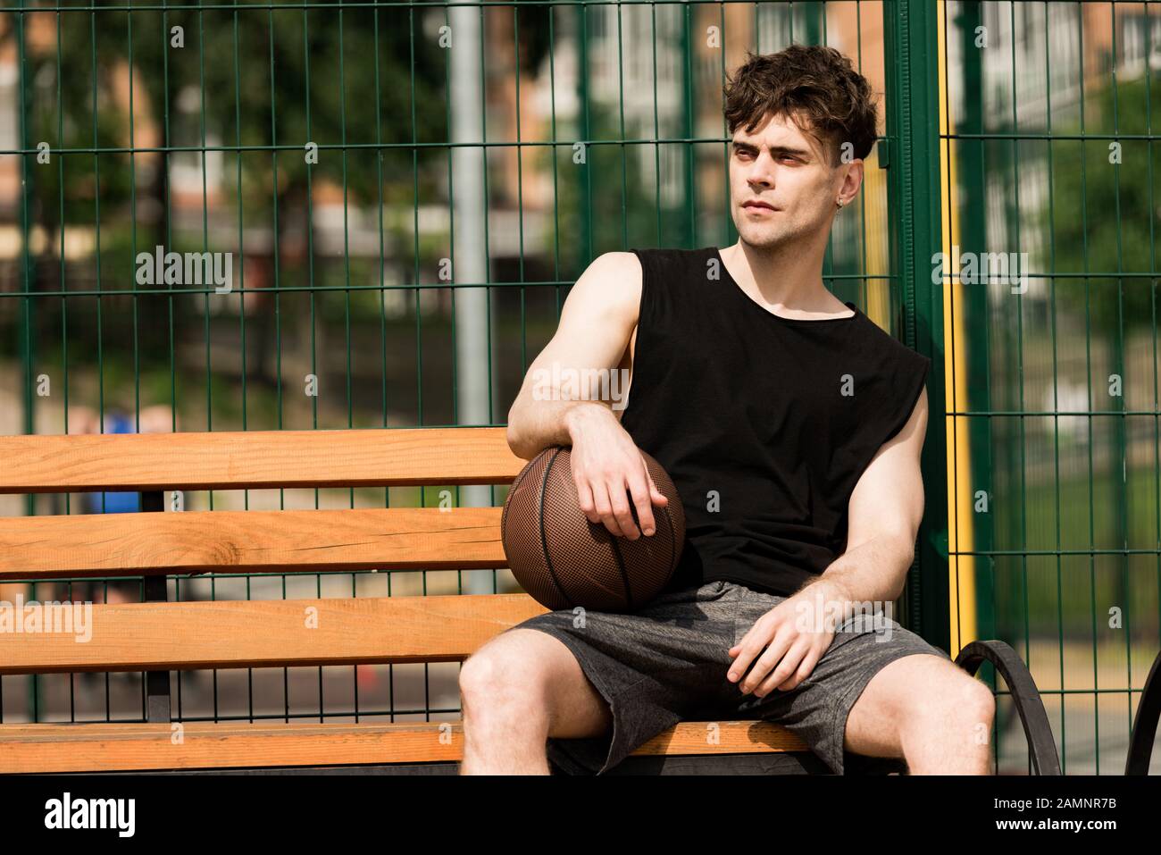 serious basketball player with ball sitting on wooden bench in sunny ...
