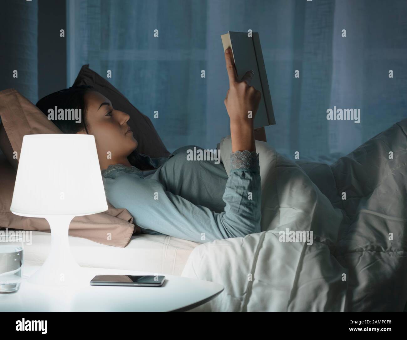 Young beautiful woman lying in bed and reading a book late at night ...