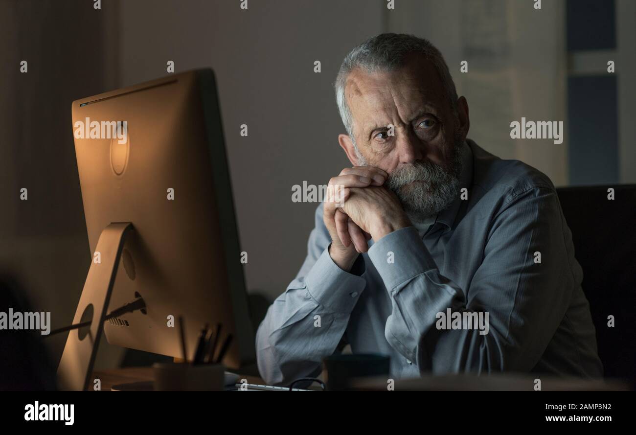 Senior man sitting at desk and working with his computer late at night ...