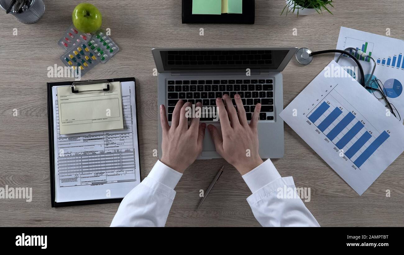 Physician looking at statistics chart, typing on laptop, preparing ...