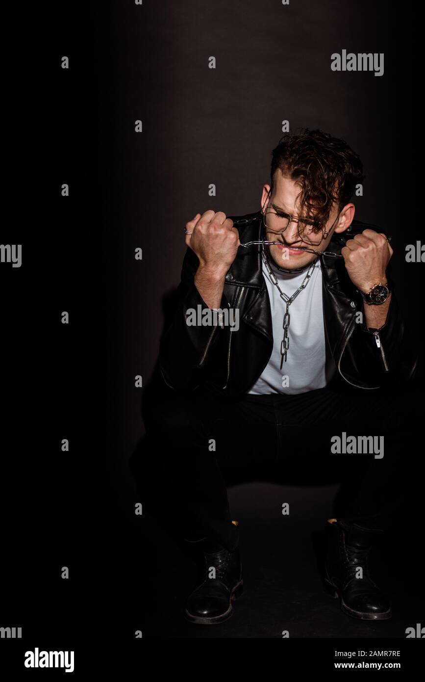 emotional man in glasses and leather jacket biting chain on black Stock ...