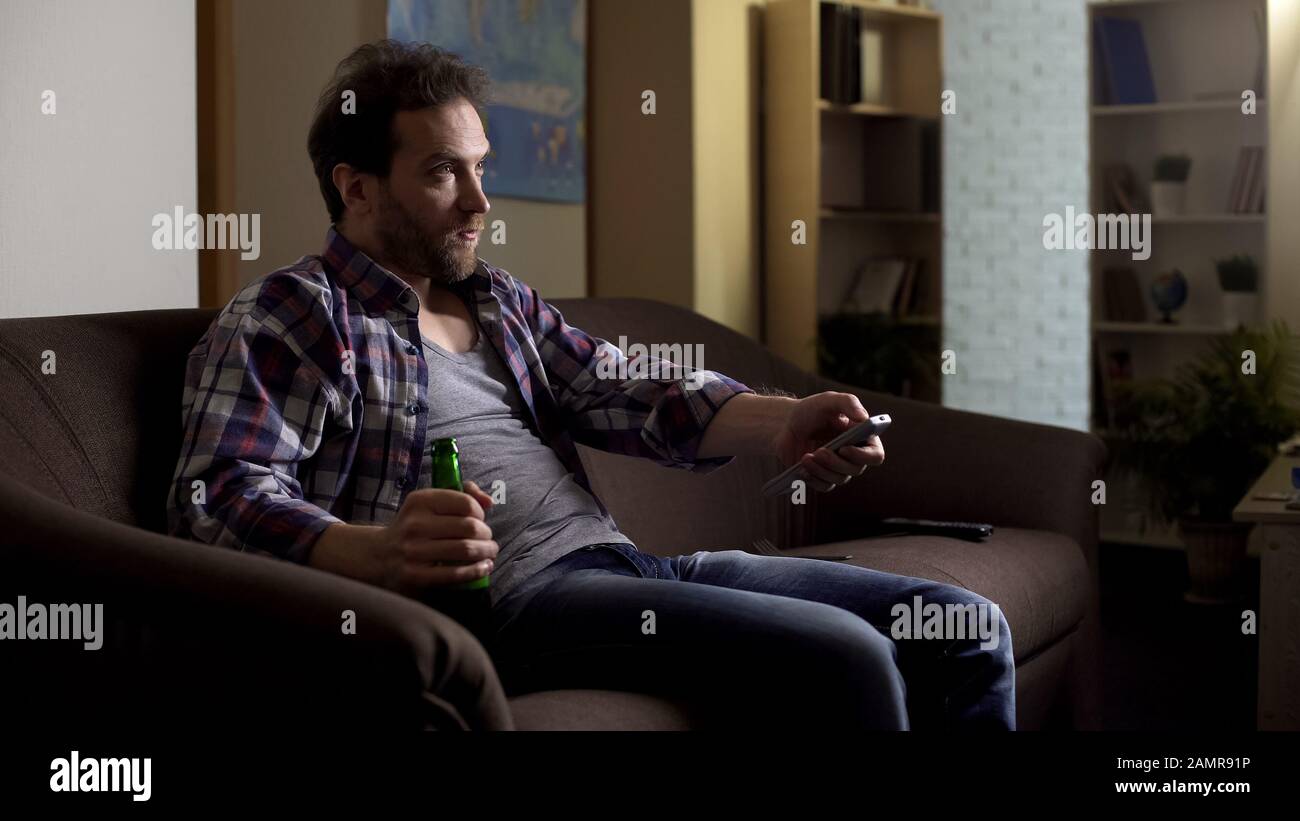 Guy sitting on sofa with beer bottle in hand, using remote control to ...
