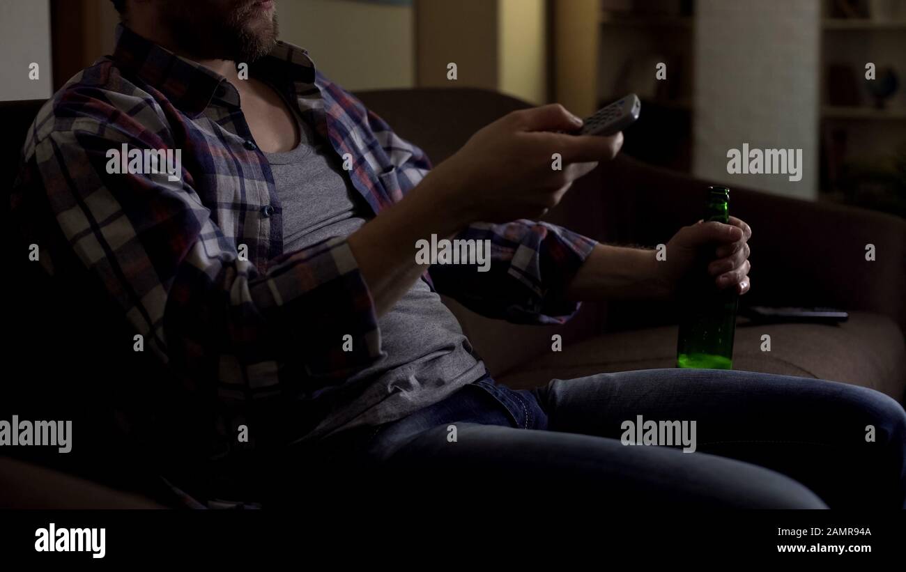 Male holding bottle of beer on knee, switching TV channels with remote ...