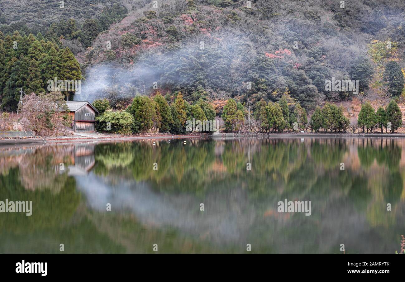 Hut with chimney smoke over Lake Suigetsu and reflected in the water ...