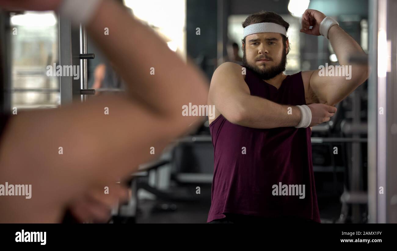 Frustrated overweight man looking at his hand fat dreaming about biceps ...