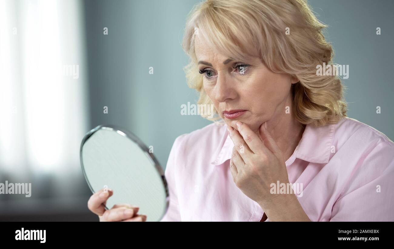 Unhappy senior female looking at sagging skin face in mirror, old age ...