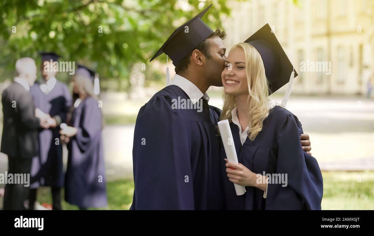 Graduates in academic regalia smiling, happy guy kissing girlfriend on ...