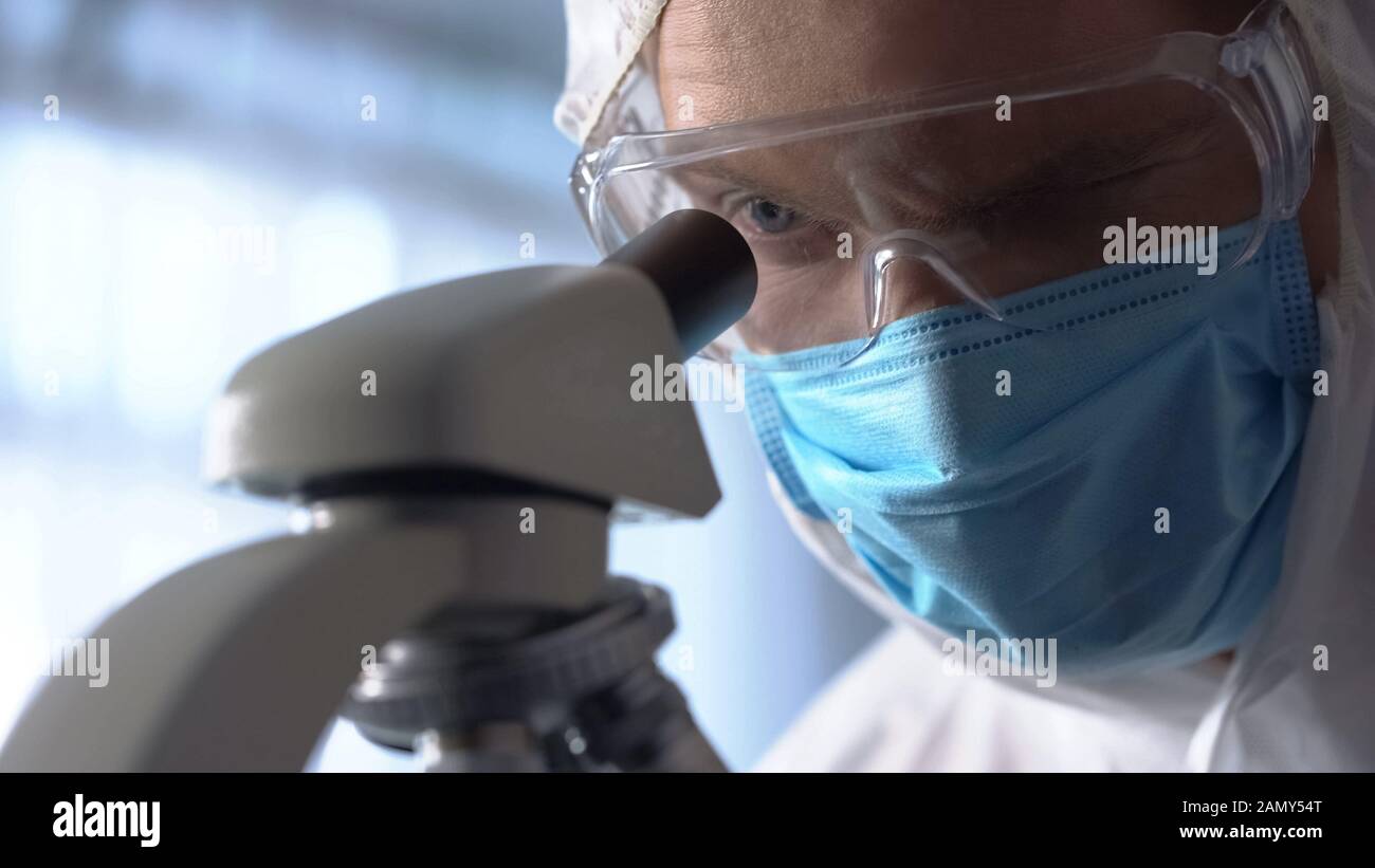 Male lab assistant in protective mask and eyewear using microscope for ...