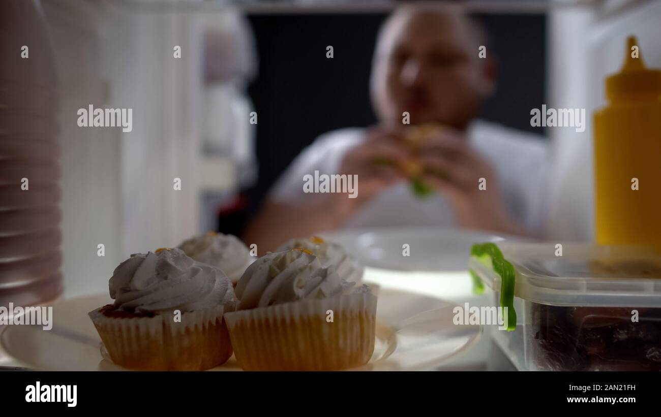 Fat man eating burger in front of fridge, junk food addiction, cupcakes ...