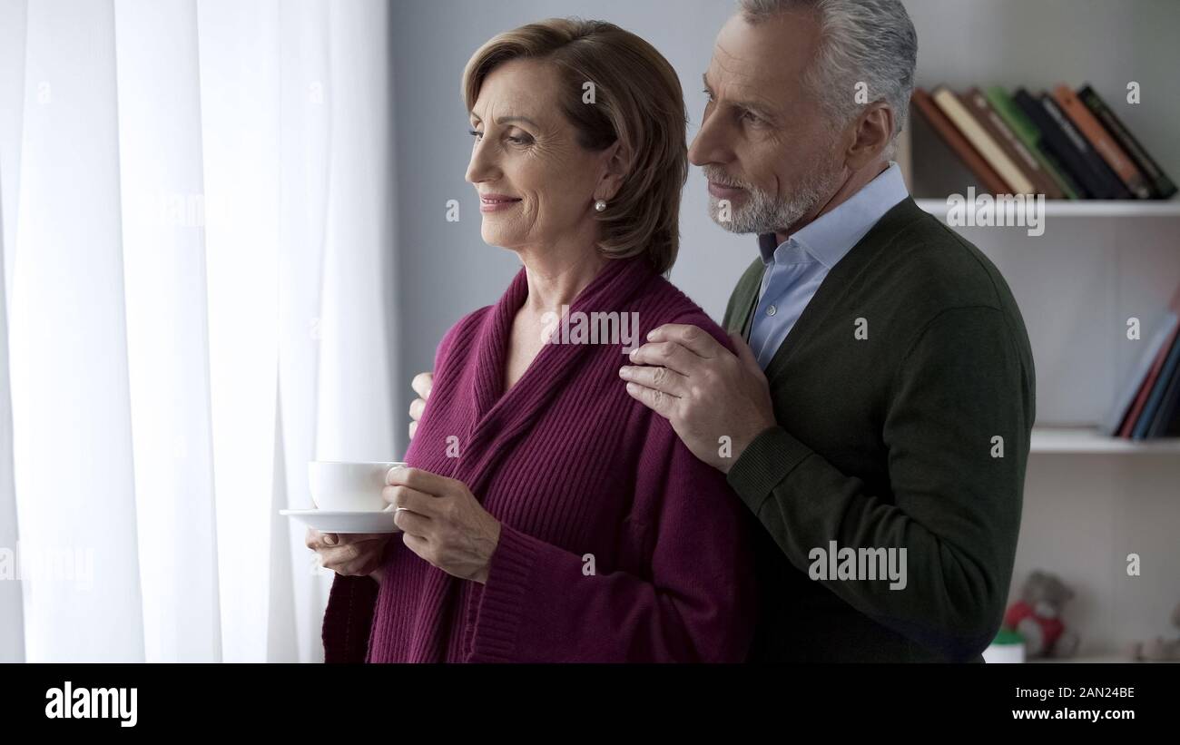 Senior couple looking in window, male hugging lady shoulders from ...