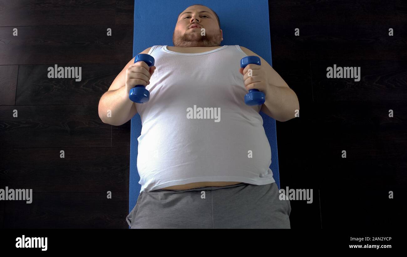 Purposeful fat man lifting dumbbells with great effort lying on mat ...