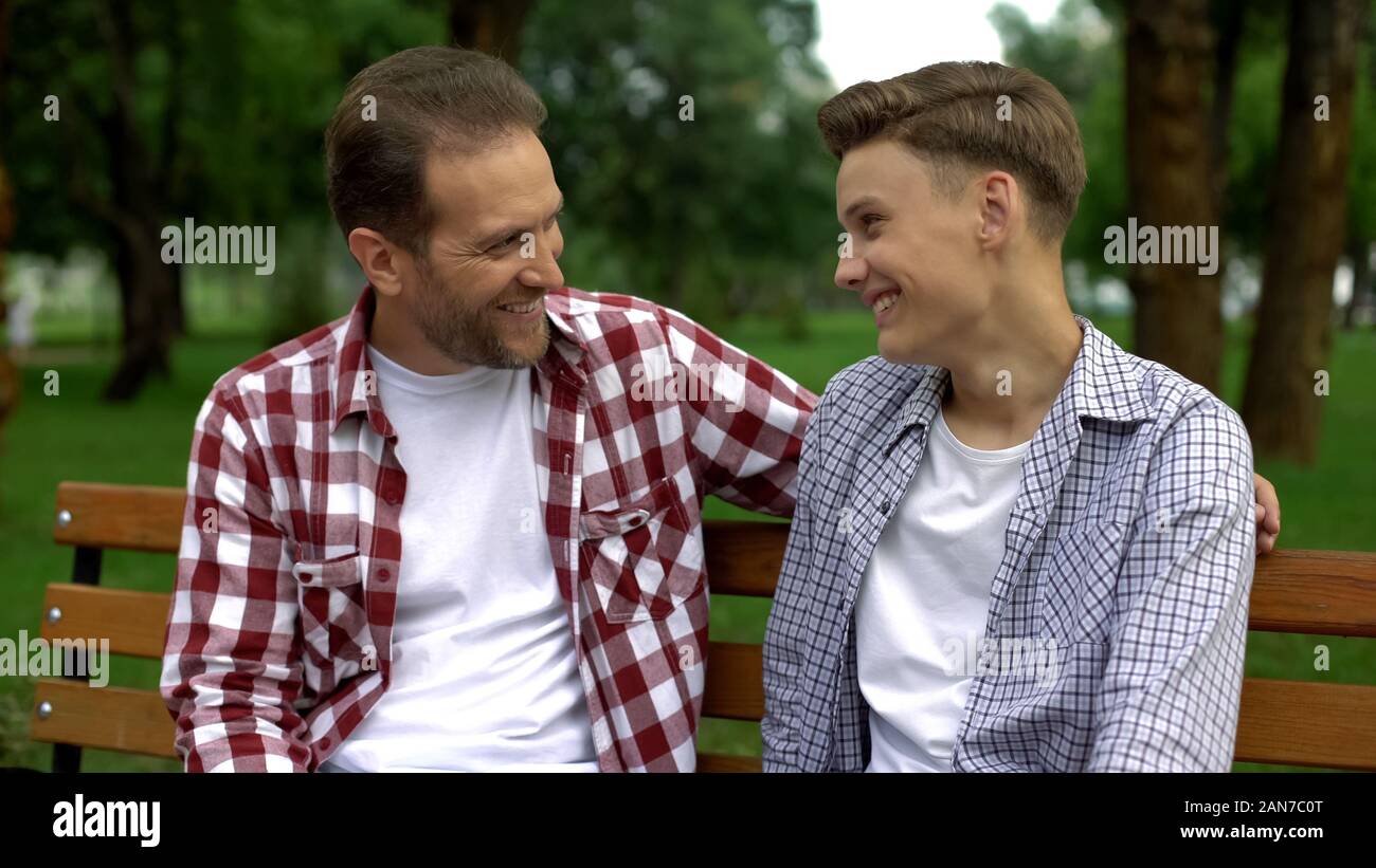 Teenage son talking with father on bench, telling secrets and smiling ...