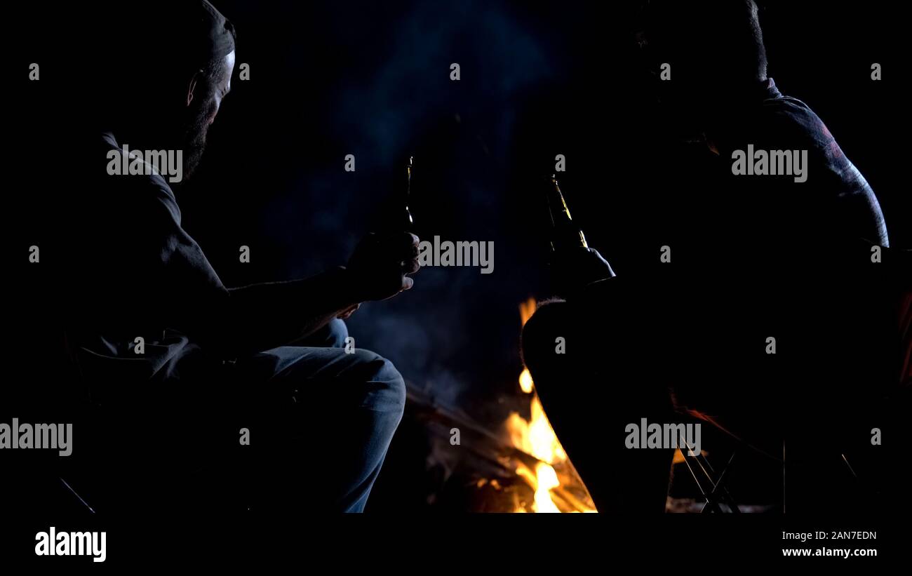 Two friends sitting around open fire at night and drinking beer ...