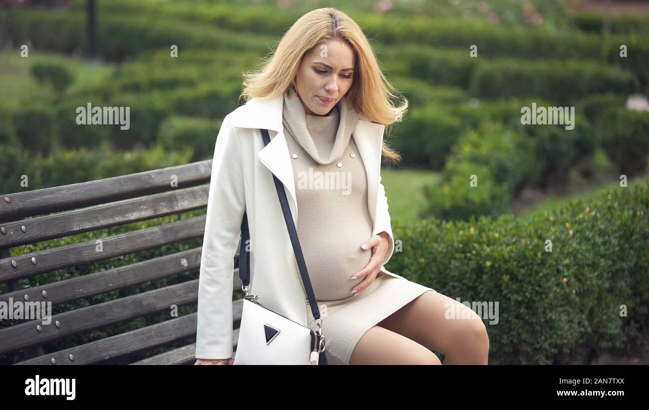 Worried expecting lady touching her belly, sitting on bench, looking ...