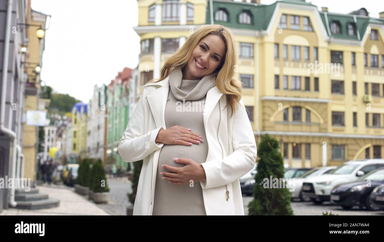 Happy expecting woman looking at camera and smiling, stroking belly ...