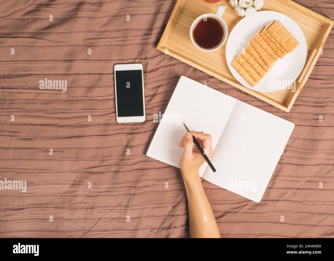 woman writes in large white open notebook, lay on bed with smartphone ...